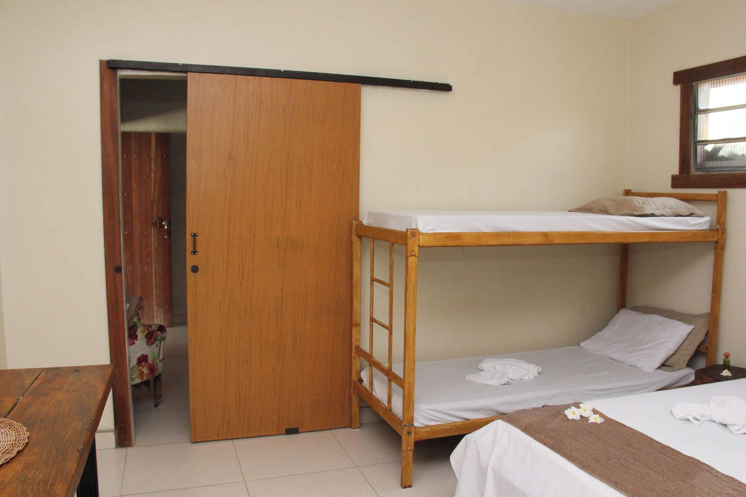Quarto individual com uma cama beliche de madeira, porta que dá para outro quarto e uma pequena mesa com um tapete de fibra natural.