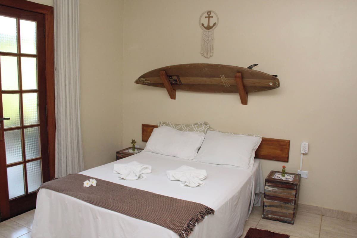 Cama bem arrumada com lençóis brancos e manta marrom, uma prancha de surf de madeira fixada na parede acima e uma pequena mesa de cabeceira de madeira com um vaso de flores ao lado da cama.