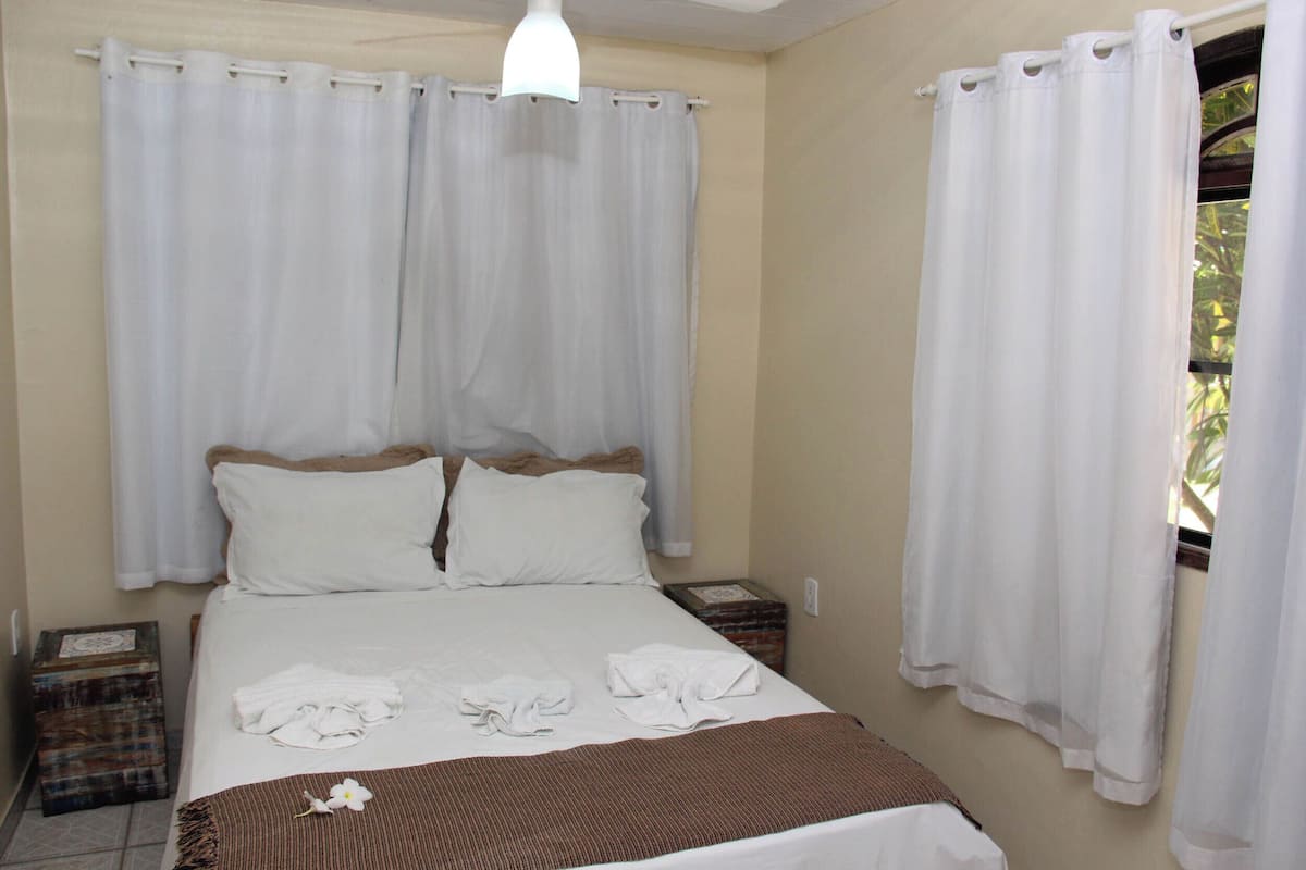 Uma cama bem arrumada com lençóis brancos e um cobertor marrom, uma mesa de cabeceira com abajur, e uma janela com cortinas brancas.