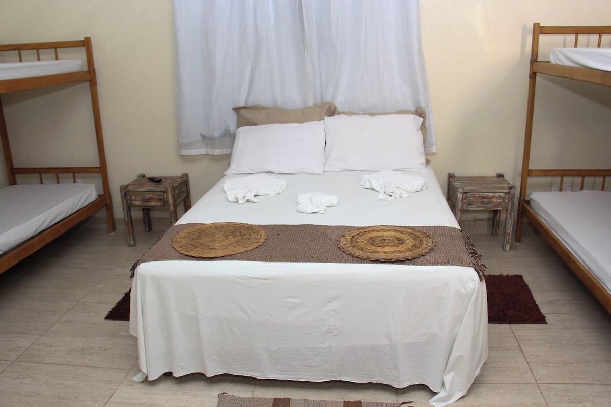 Cama de solteiro com colcha branca e dois jogos americanos trançados, com duas mesinhas de cabeceira de madeira, uma pequena mesa e uma cadeira.