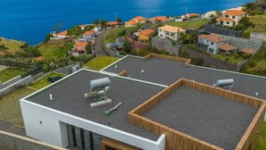Um edifício moderno com telhado plano, uma pequena área externa com churrasqueira e vista para o oceano e casas próximas.