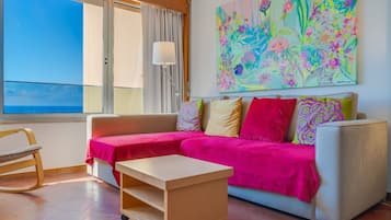 Quarto com sofá, mesa pequena, cadeira, pintura e janela com vista.