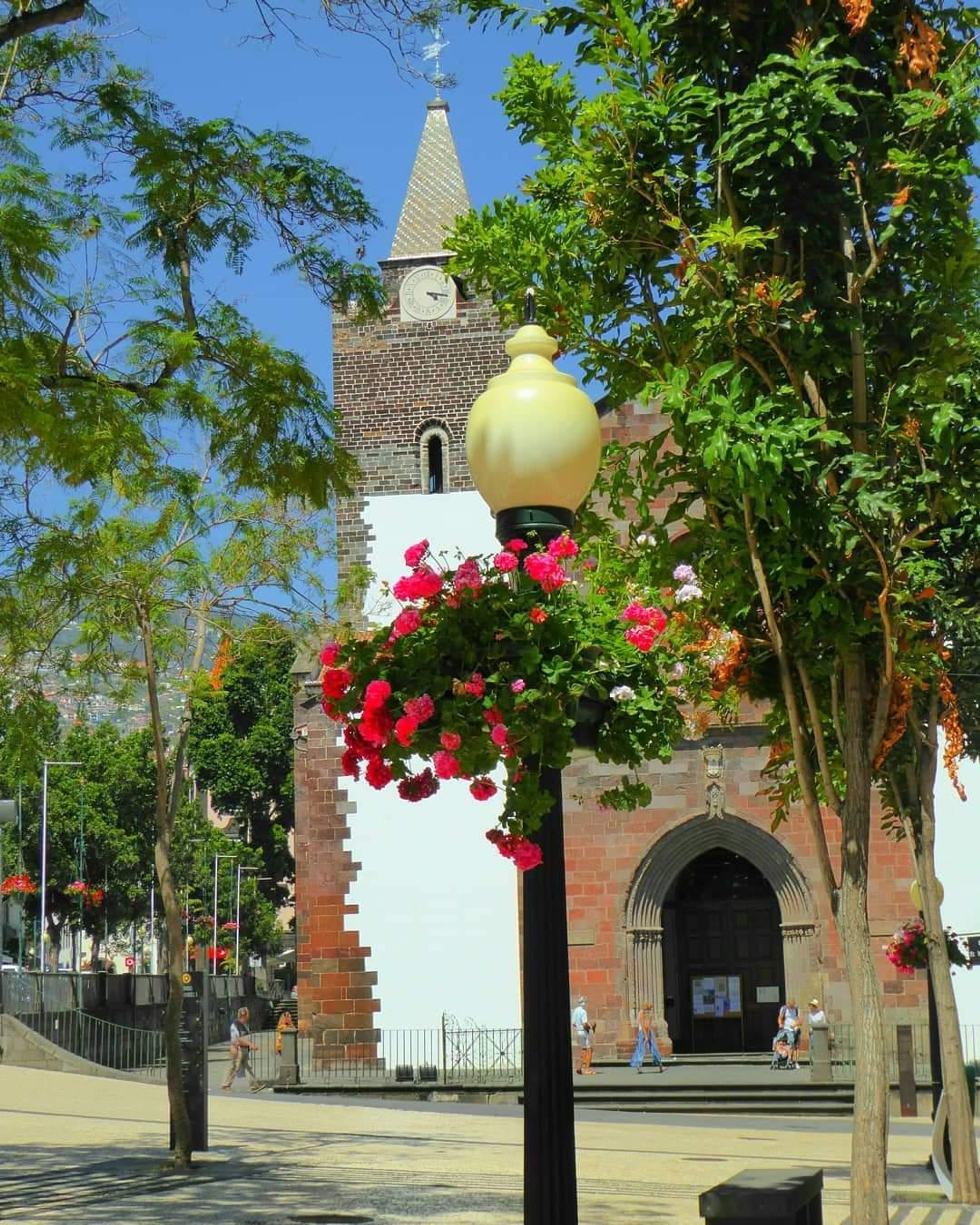 Cena de rua com um edifício histórico, uma torre de relógio, um poste de luz com flores e pedestres.