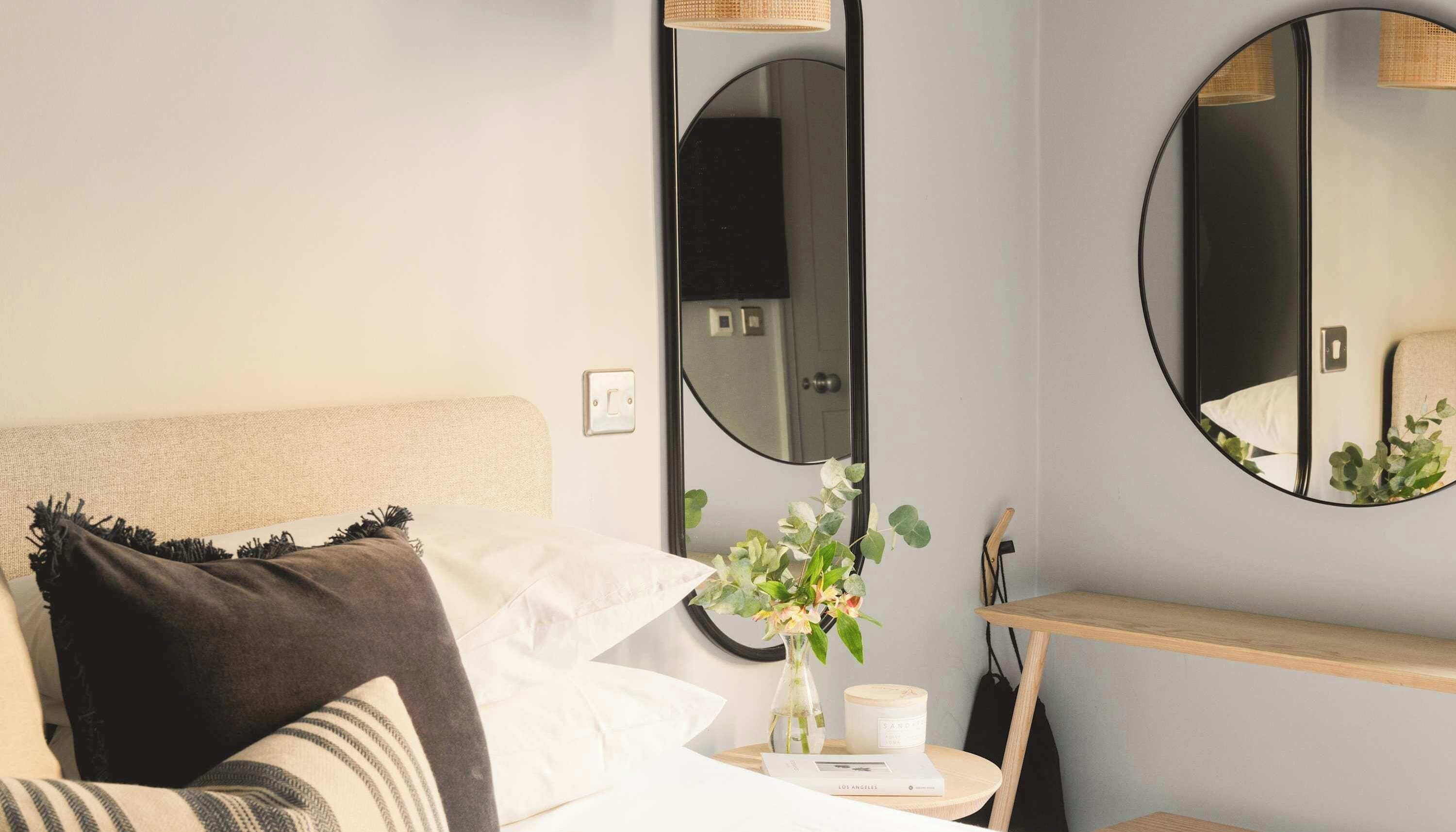Quarto de hotel com cama, mesa de cabeceira com um vaso de flores, espelho e prateleira de madeira.