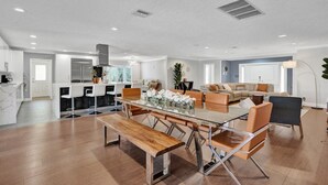 Uma sala de estar ampla com uma grande mesa de jantar de vidro, assentos confortáveis, uma área de cozinha e um espaço aconchegante com um sofá e poltronas.