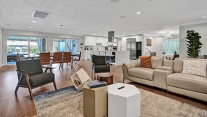 Uma sala de estar moderna com um sofá, cadeiras e uma mesa de centro. Há uma área de refeições com uma mesa e cadeiras. O aposento tem janelas amplas e vista para o exterior.