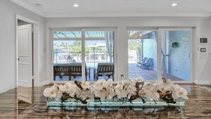 Um quarto espaçoso com uma grande mesa de vidro ao centro, vista para um pátio externo com assentos, e uma porta que dá para outra área.