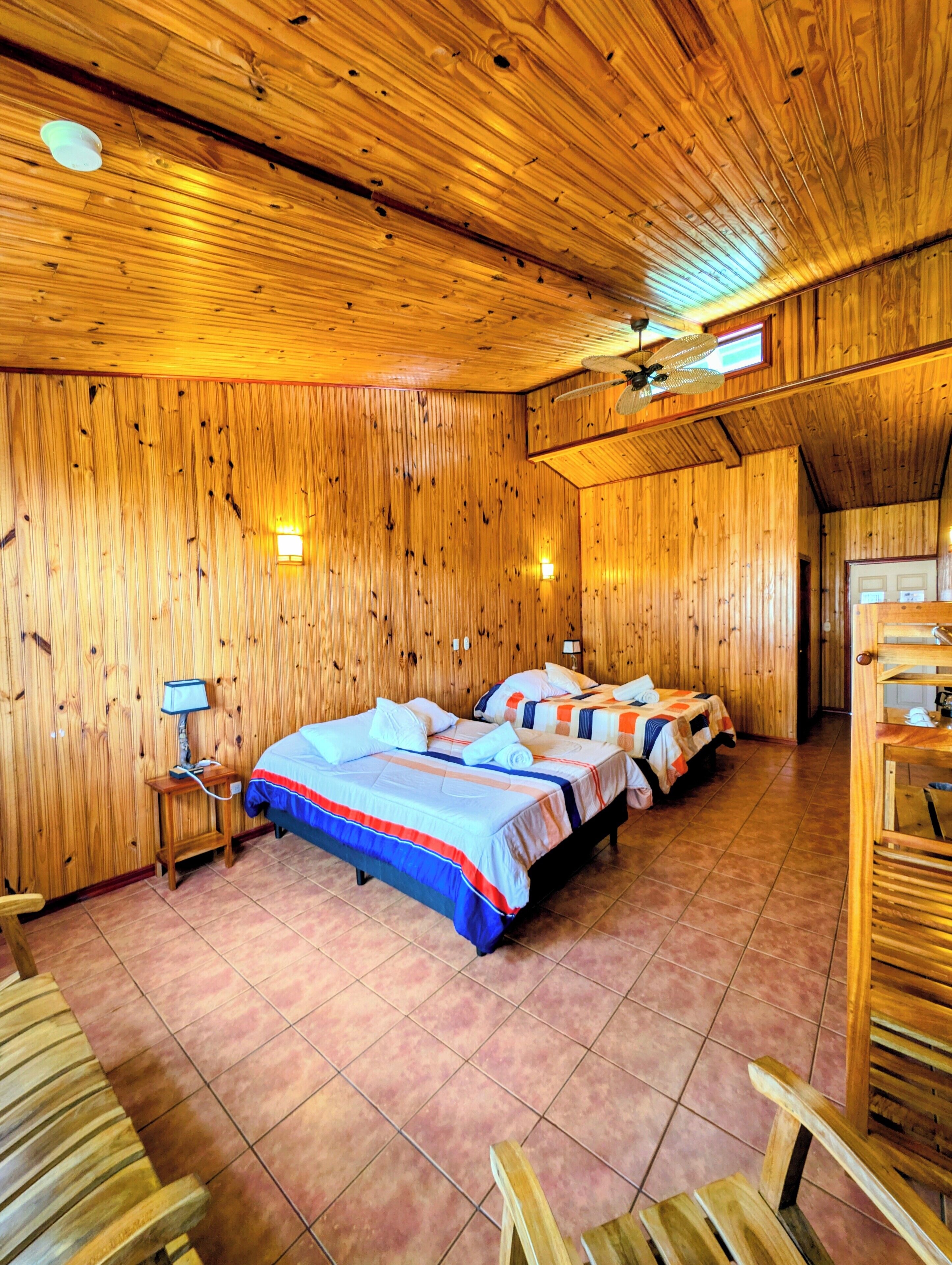 Quarto com painéis de madeira, duas camas, um ventilador de teto e uma pequena mesa com abajur.