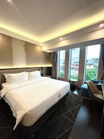 Quarto de hotel moderno com uma cama grande, uma escrivaninha pequena e vista para a cidade através de amplas janelas.