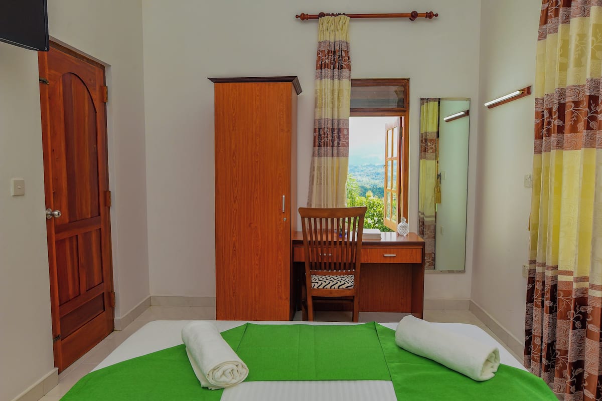 Quarto com cama, escrivaninha, cadeira, guarda-roupa, espelho e janela com vista.