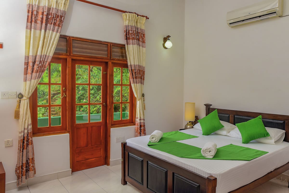 Cama bem arrumada com almofadas verdes e toalha enrolada, porta de madeira com janela e ar-condicionado fixado na parede.