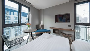 Quarto de hotel com uma cama, uma mesinha com um vaso, uma cadeira, um banco e uma TV fixada na parede.