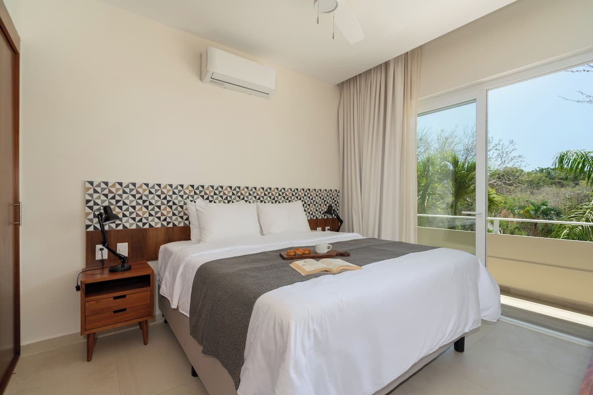 Quarto de hotel com cama, mesa de cabeceira, luminária para leitura, janela com vista para o verde e ventilador de teto.