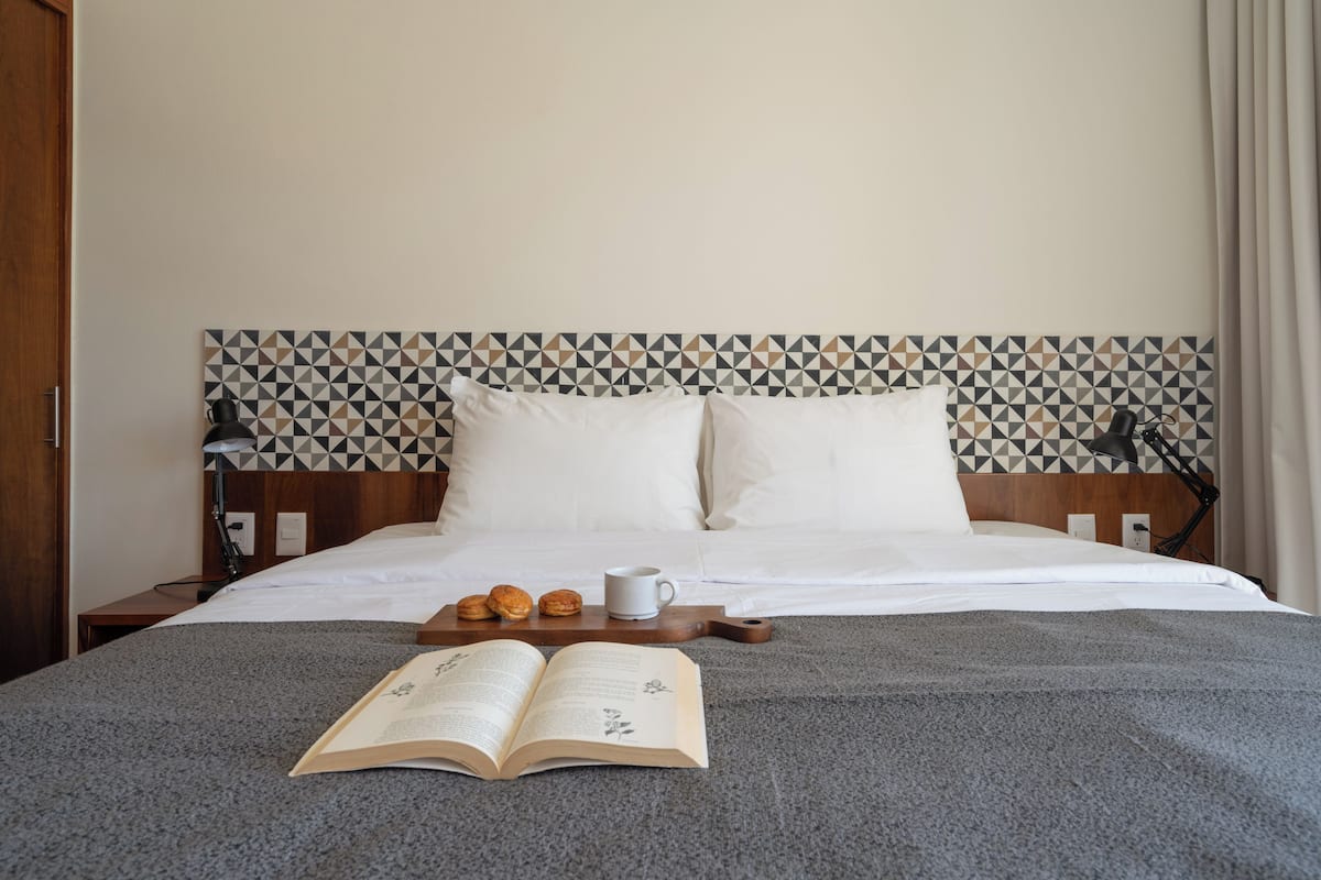 Uma cama bem arrumada com cabeceira estampada, mesa de cabeceira com abajur e bandeja com doces e uma xícara.