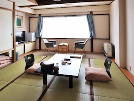 Quarto com esteiras de tatame, uma mesa baixa com almofadas, uma televisão e uma janela grande com cortinas.