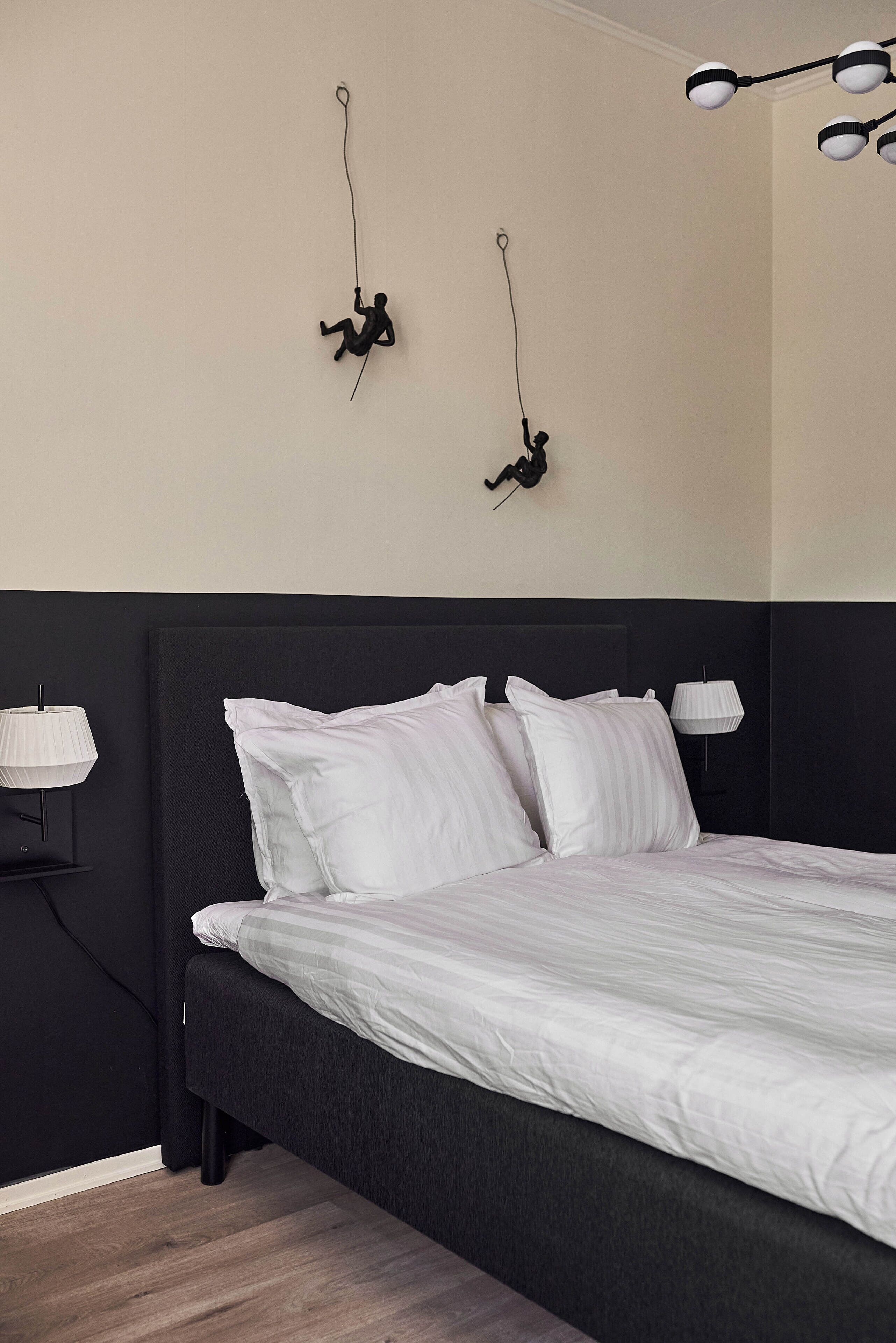 Cama bem arrumada com lençóis brancos, cabeceira escura e duas luminárias fixadas na parede.