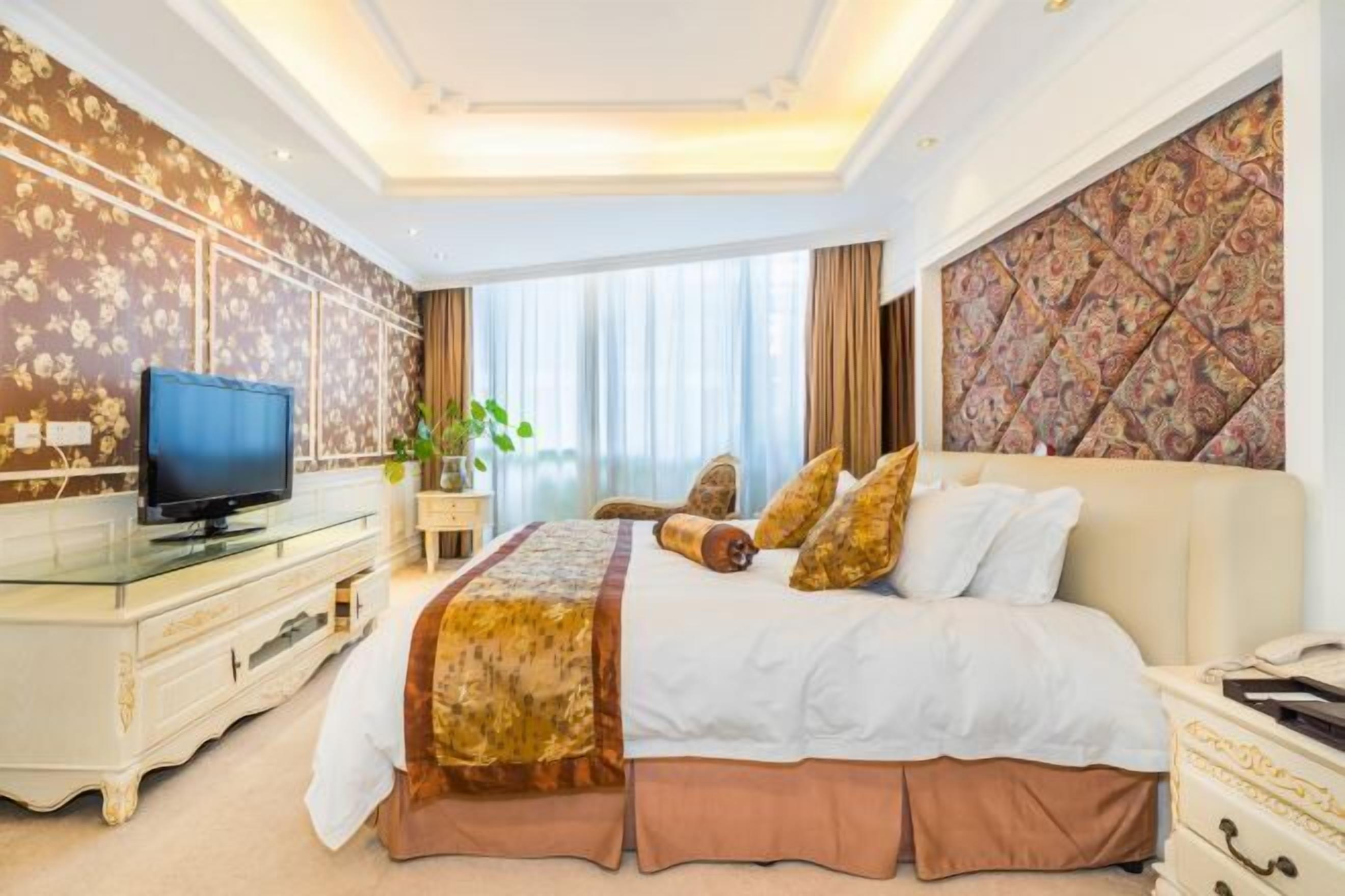 Quarto de hotel com uma cama grande, televisão, uma planta e uma cabeceira decorativa.