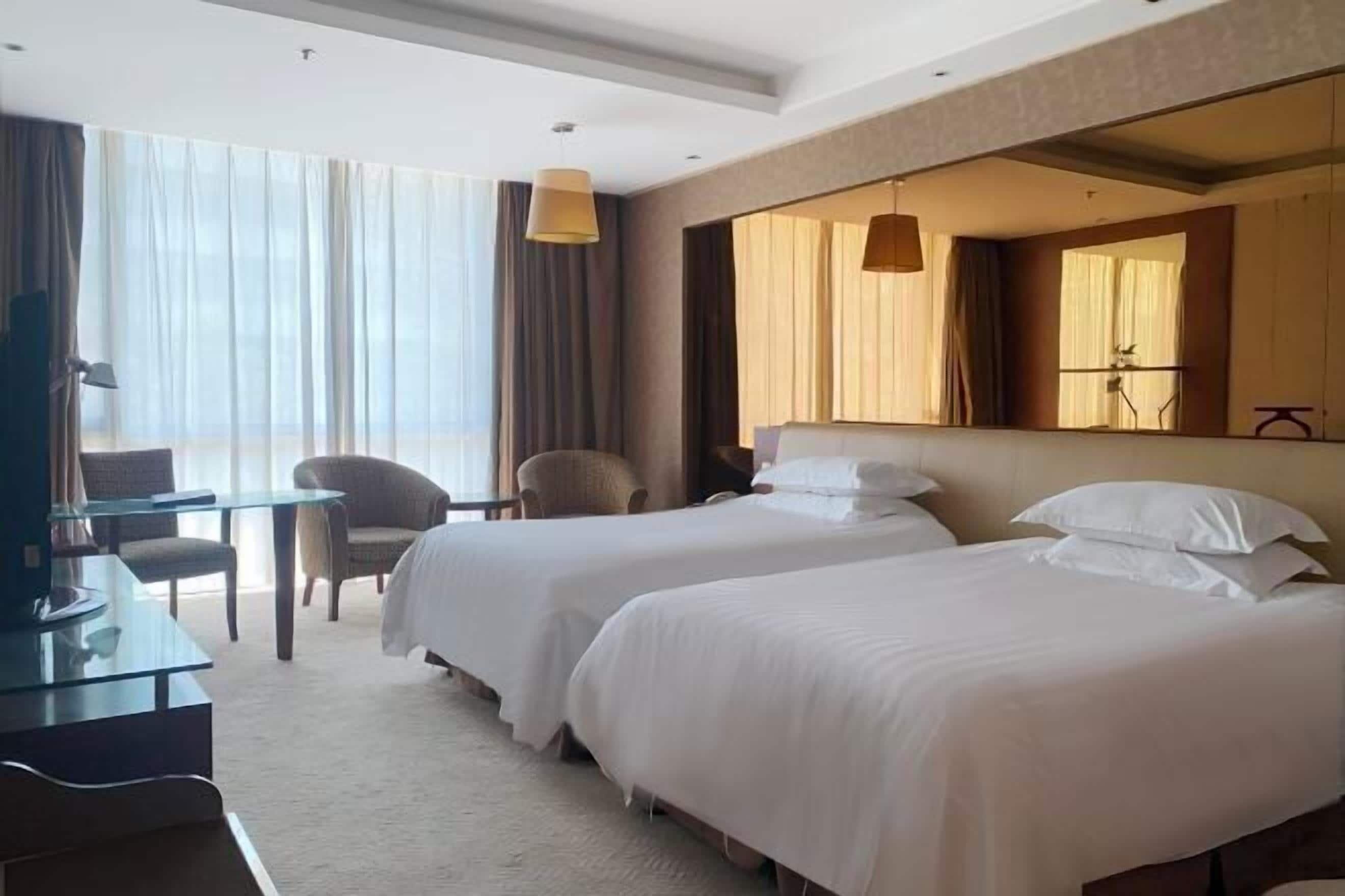 Quarto de hotel com duas camas, uma escrivaninha, uma cadeira e uma janela grande com cortinas.