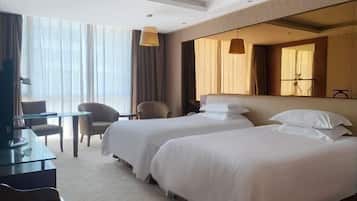Quarto de hotel com duas camas, uma escrivaninha, uma cadeira e uma janela grande com cortinas.
