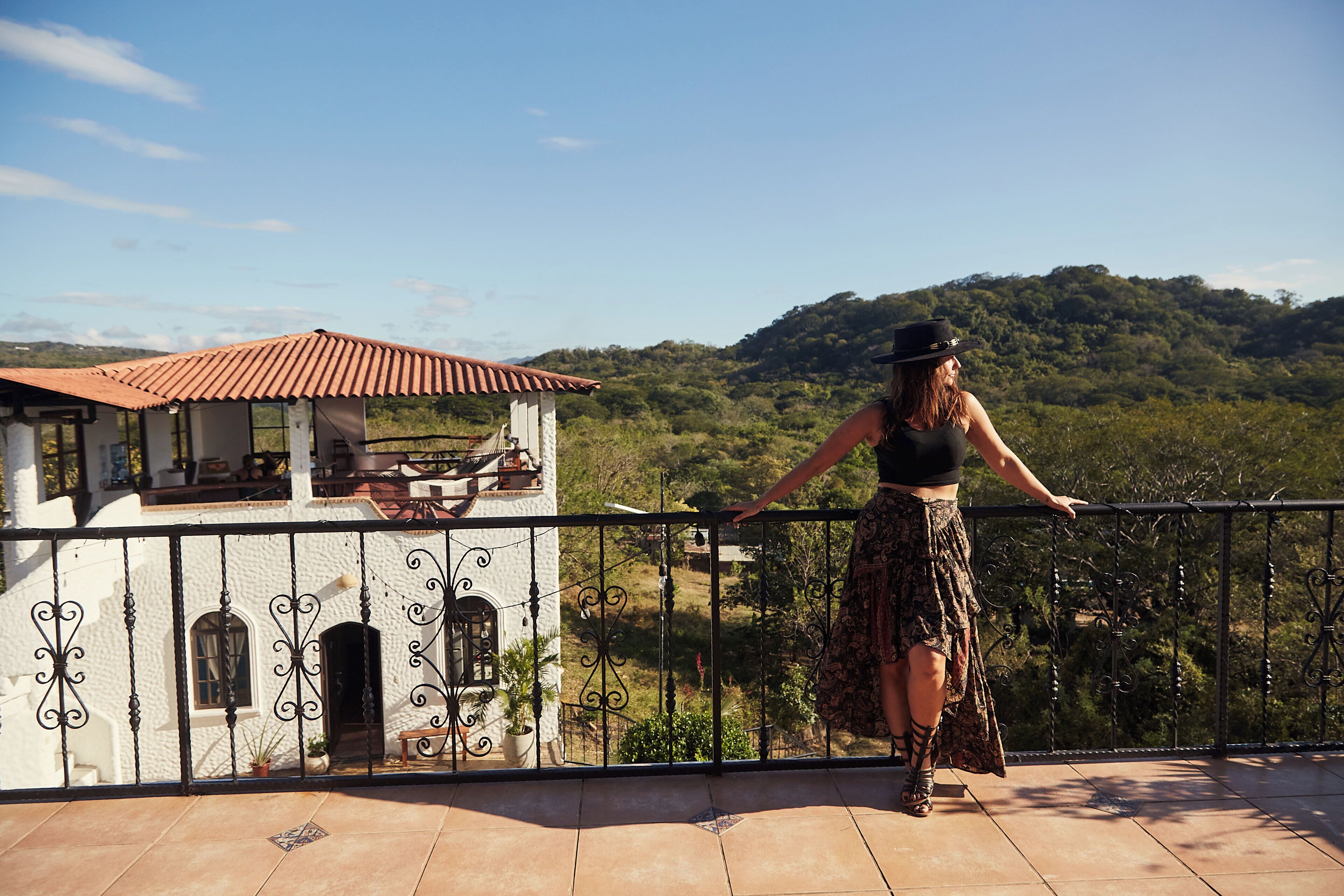 Varanda com vista para um edifício branco com telhado vermelho e guarda-corpo preto, e uma mulher em pé na varanda.