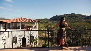 Varanda com vista para um edifício branco com telhado vermelho e guarda-corpo preto, e uma mulher em pé na varanda.