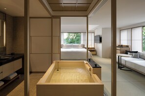 Quarto de hotel moderno com uma grande banheira de madeira, um quarto separado com uma cama e uma área de estar com um sofá e uma pequena mesa.