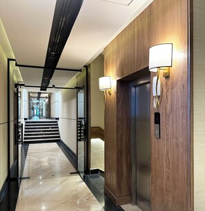 Corredor moderno de hotel com piso de mármore, painéis de madeira e escada que leva a um elevador.