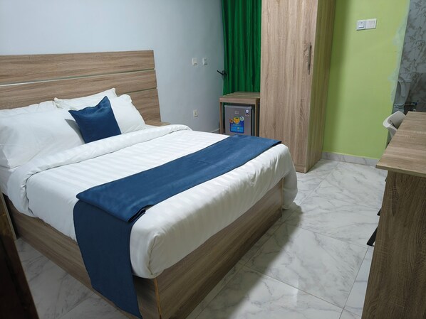 Cama bem arrumada com roupa de cama branca e azul, uma pequena mesa de cabeceira com uma garrafa e uma cortina verde num quarto com paredes claras e cabeceira de madeira.