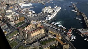 Vista aérea de uma cidade costeira com um porto importante, edifícios históricos e um grande navio de cruzeiro atracado.