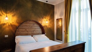 Quarto com cabeceira de madeira, duas luminárias de apoio, cama com roupa de cama branca, mesa de cabeceira de madeira e janela com cortinas translúcidas.