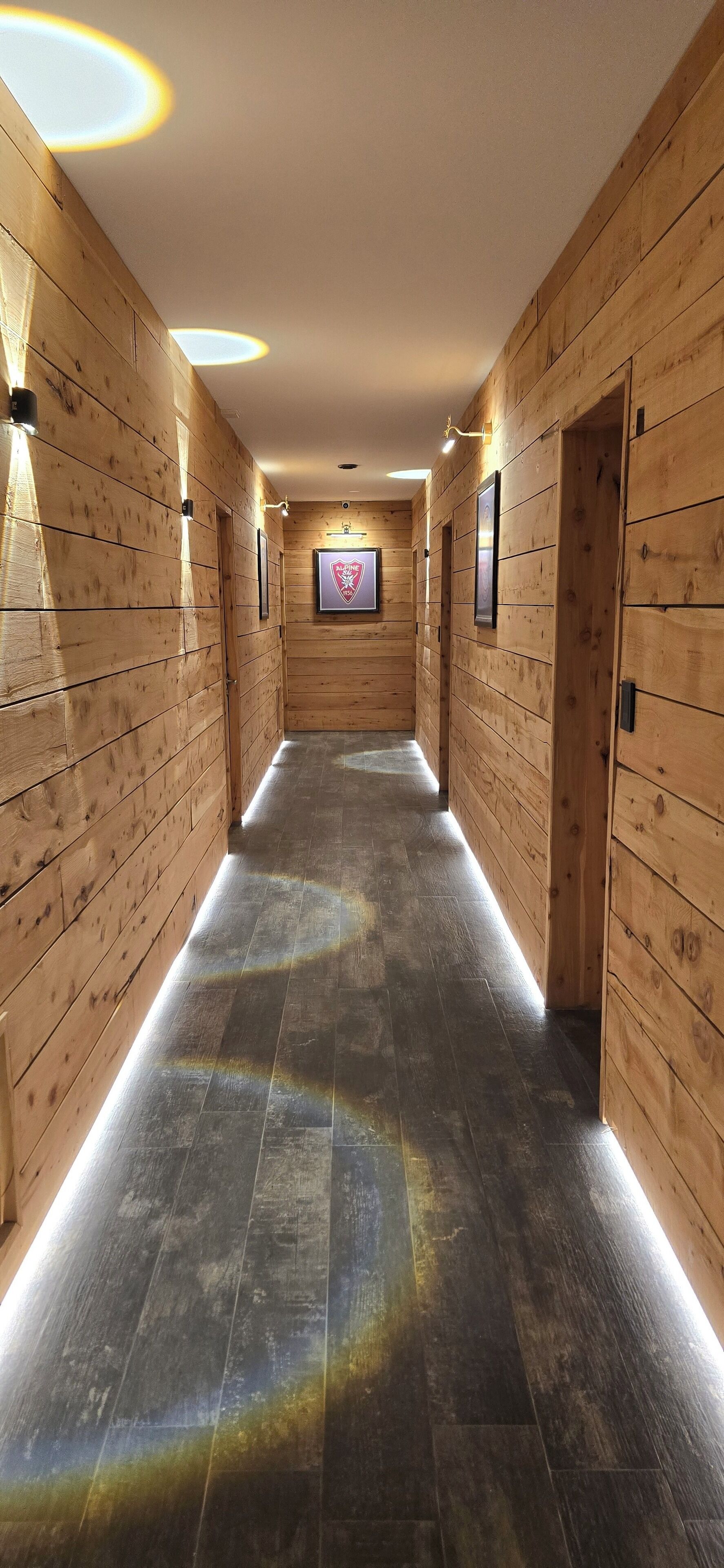 Um corredor revestido de madeira com iluminação embutida, um brasão emoldurado na parede e uma porta à direita.