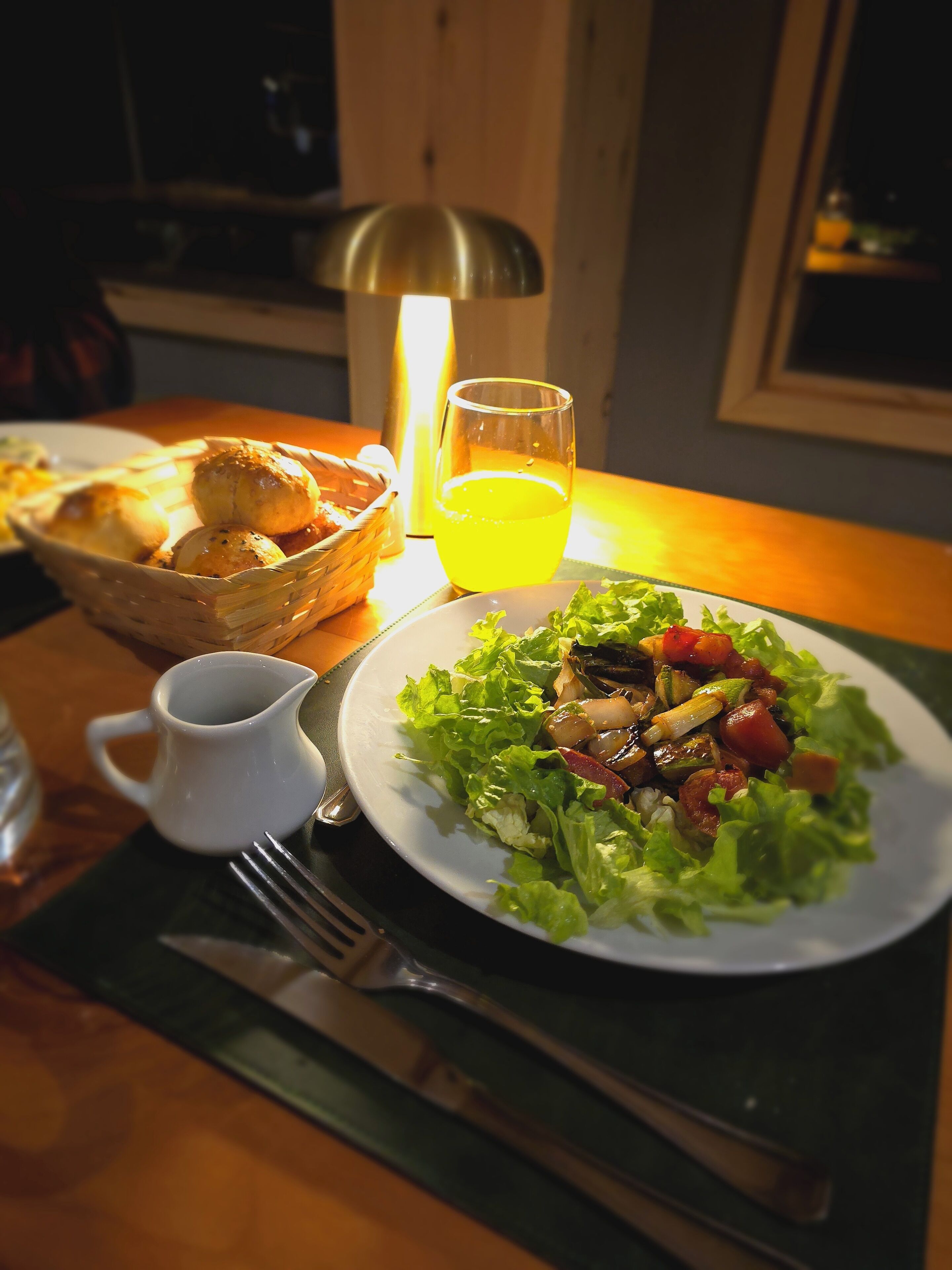 Uma mesa com uma salada, um copo de suco de laranja, uma cesta de pães e uma pequena jarra.