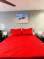 Uma cama com colcha vermelha, dois travesseiros vermelhos, um quadro com barcos numa praia e duas luminárias de cabeceira.