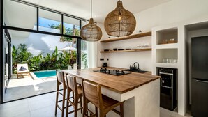 Uma cozinha moderna com uma ilha grande, banquetas, luminárias pendentes de vime e nichos embutidos, e vista para uma piscina e vegetação lá fora.