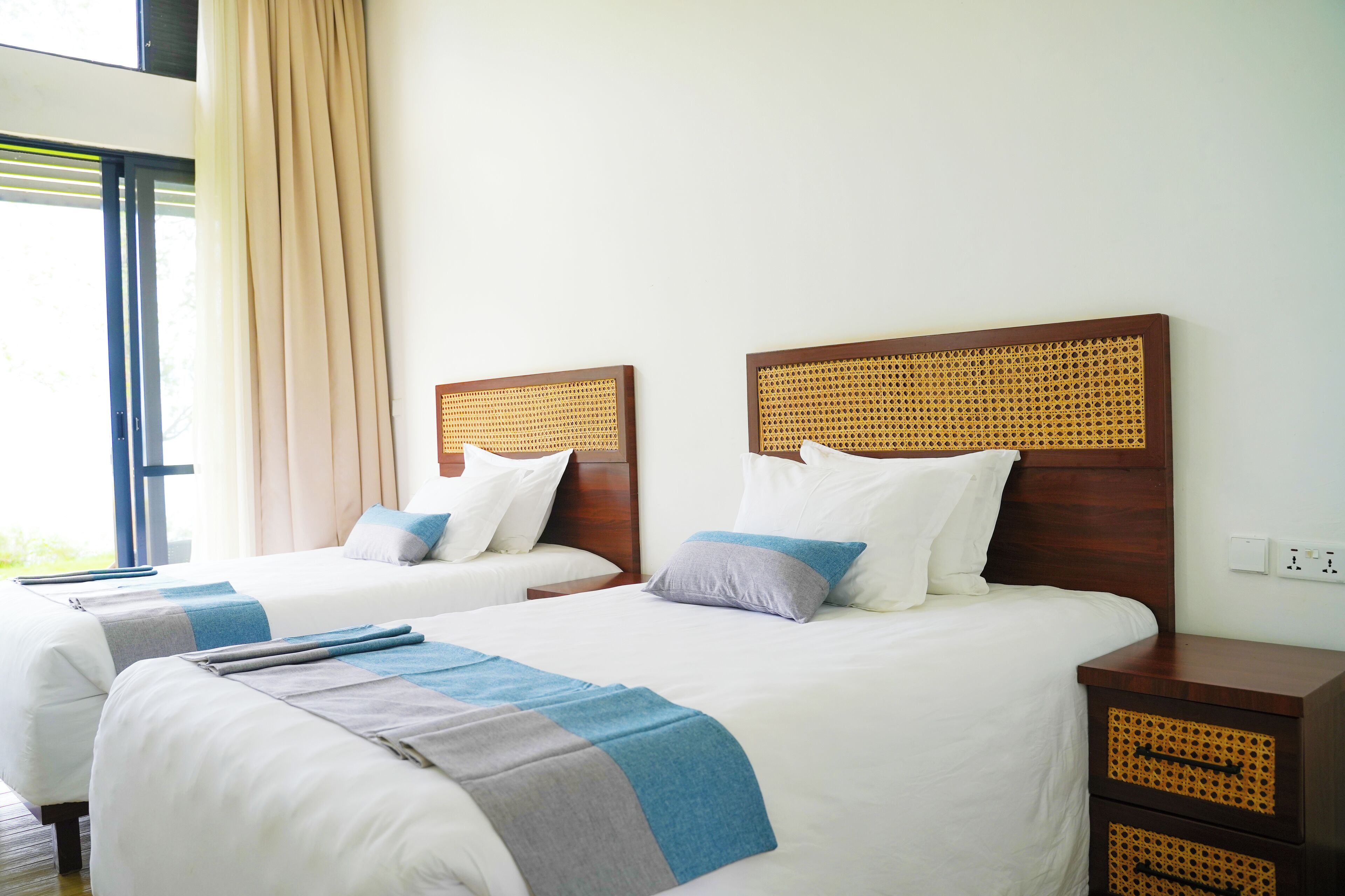 Quarto de hotel com duas camas, cada uma com cabeceira de madeira, roupa de cama branca e manta azul e cinza. Há uma mesinha de cabeceira com gavetas e uma luminária.