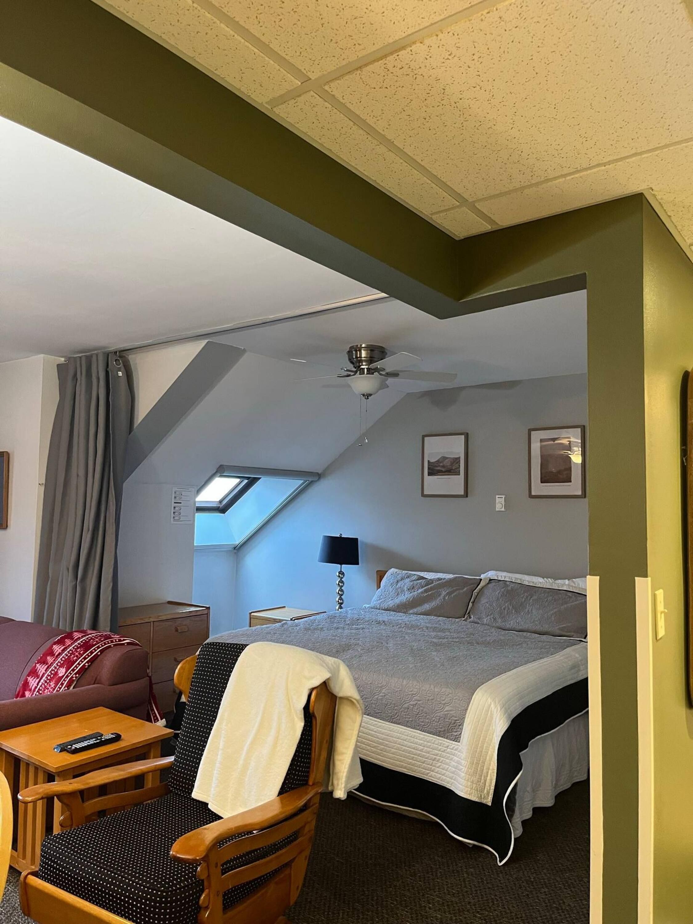 Quarto de hotel com cama, poltrona, escrivaninha, abajur, ventilador de teto e janela.