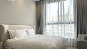 Quarto de hotel moderno com uma cama grande, uma mesinha de cabeceira e vista para a cidade através de cortinas leves.