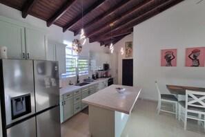 Cozinha moderna com armários brancos, eletrodomésticos de aço inoxidável e uma ilha central. Há uma área de refeições com mesa e cadeiras brancas.