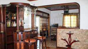 Uma área de bar com mobiliário de madeira, parede de pedra e vista para o exterior através de uma janela com cortinas.