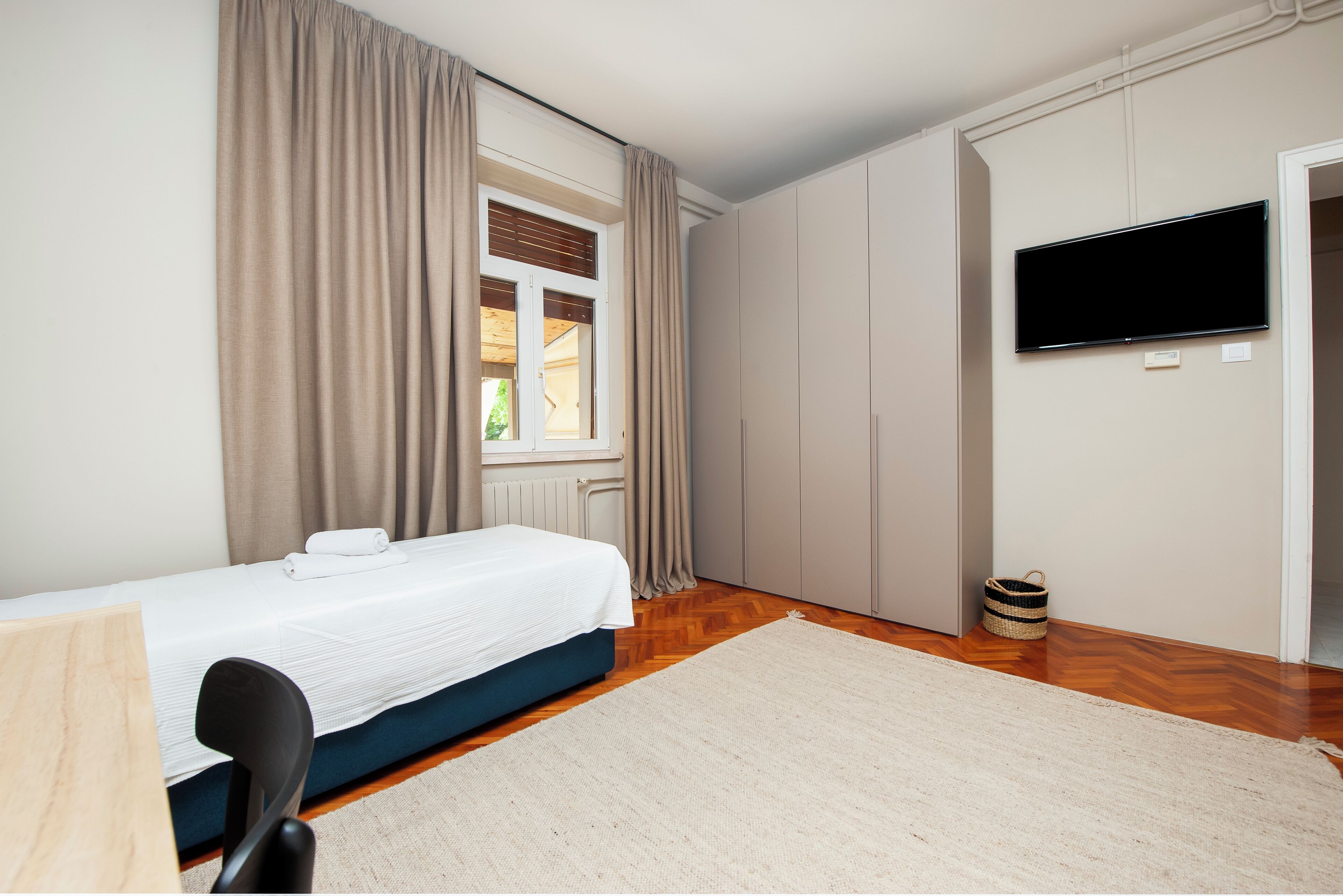 Um quarto com uma cama, escrivaninha, cadeira, televisão e janela com cortinas.