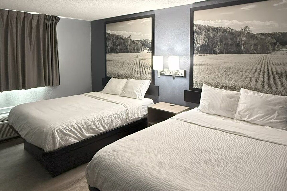 Quarto de hotel com duas camas, uma janela grande com cortinas e duas luminárias fixadas na parede.