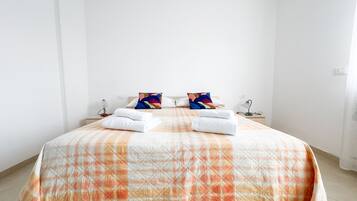 Quarto de hotel com uma cama, duas almofadas, uma mesa de cabeceira com abajur e uma tomada na parede.