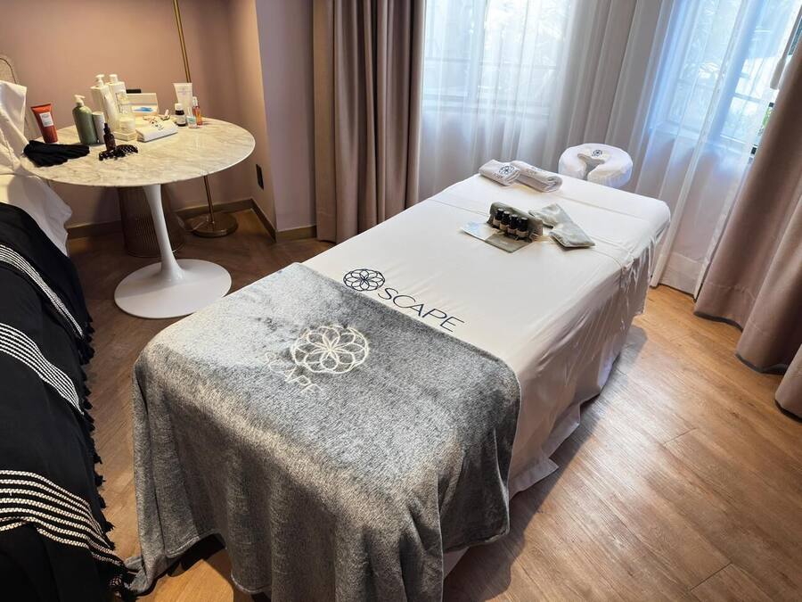 Quarto com mesa de massagem, pequena mesa redonda com artigos de higiene e cama com manta cinzenta.