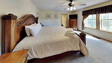 Quarto com cama grande, mesa de cabeceira de madeira, cômoda, ventilador de teto e janela com cortinas.