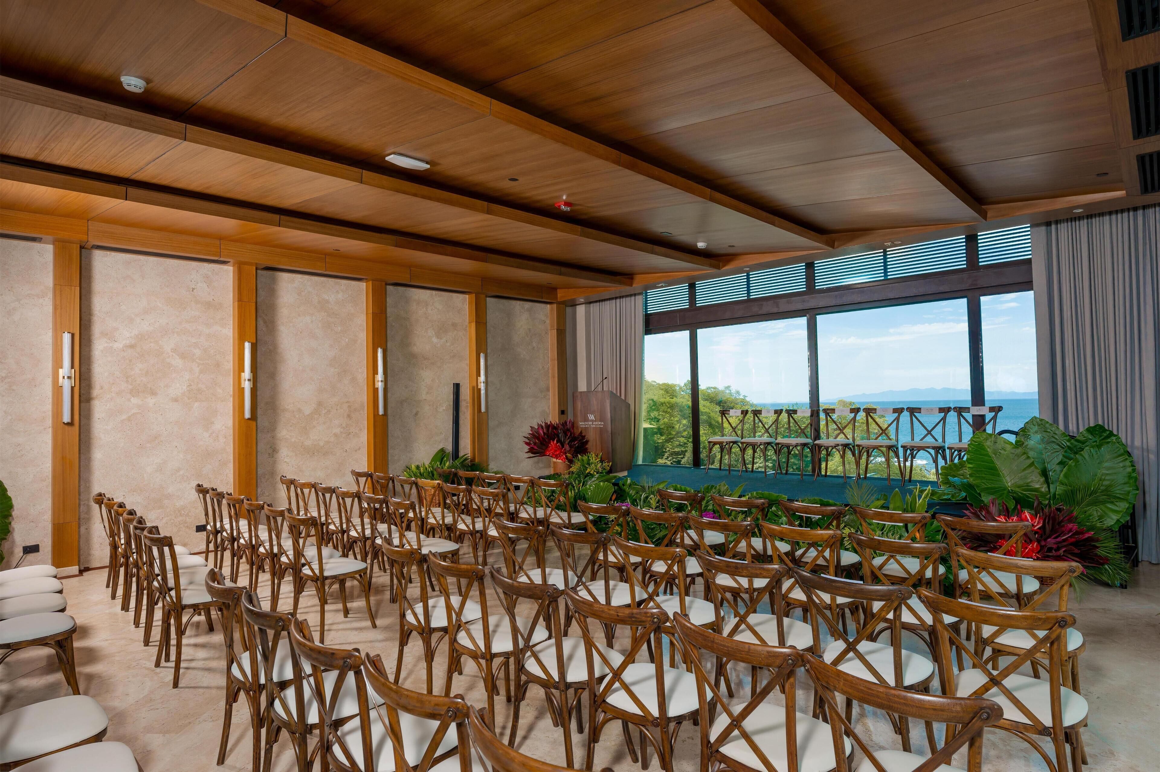 Uma sala de conferências com cadeiras e mesas de madeira, janelas amplas com vista e teto de madeira.