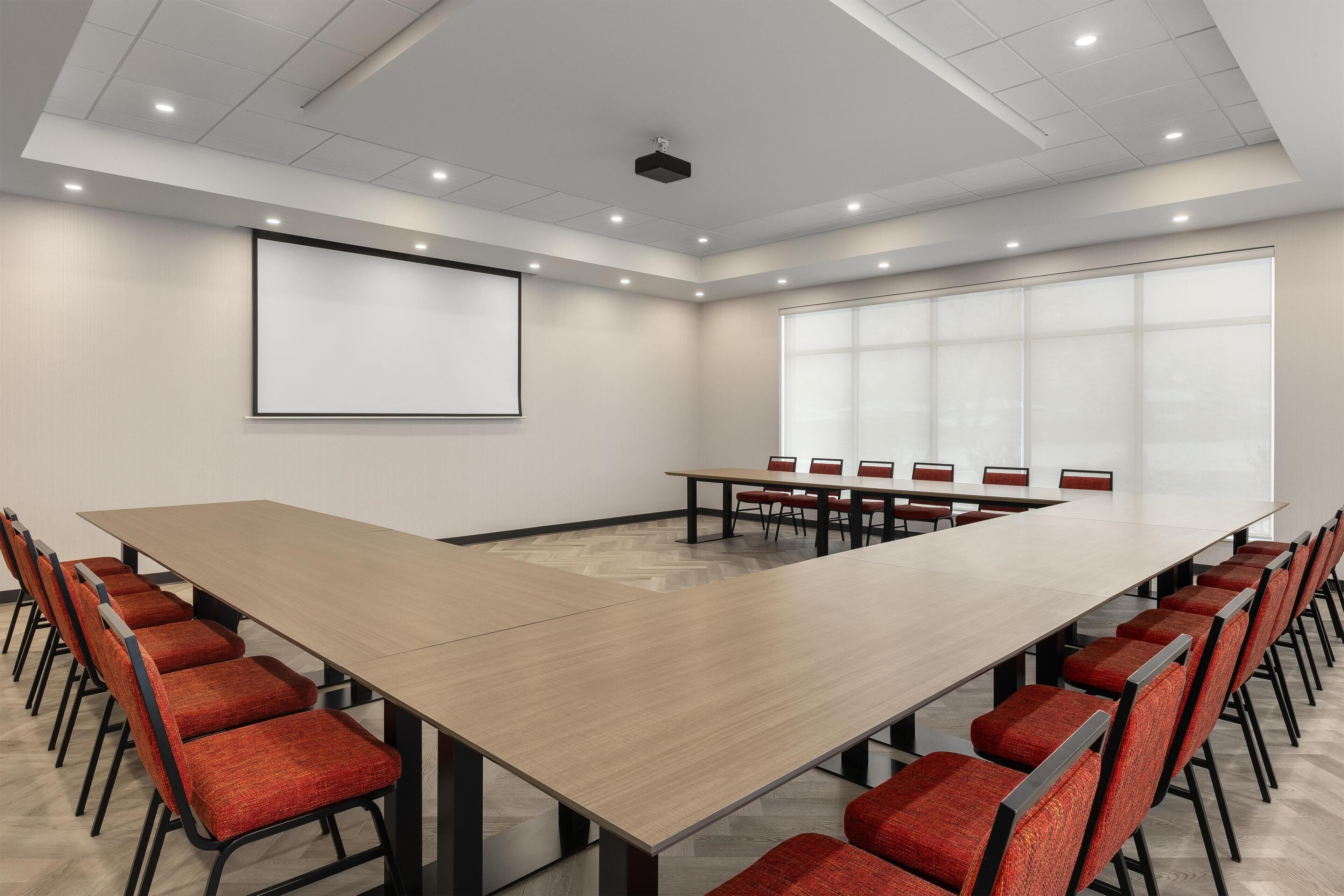 Sala de conferência com uma mesa comprida, cadeiras vermelhas, tela de projetor e grandes janelas.