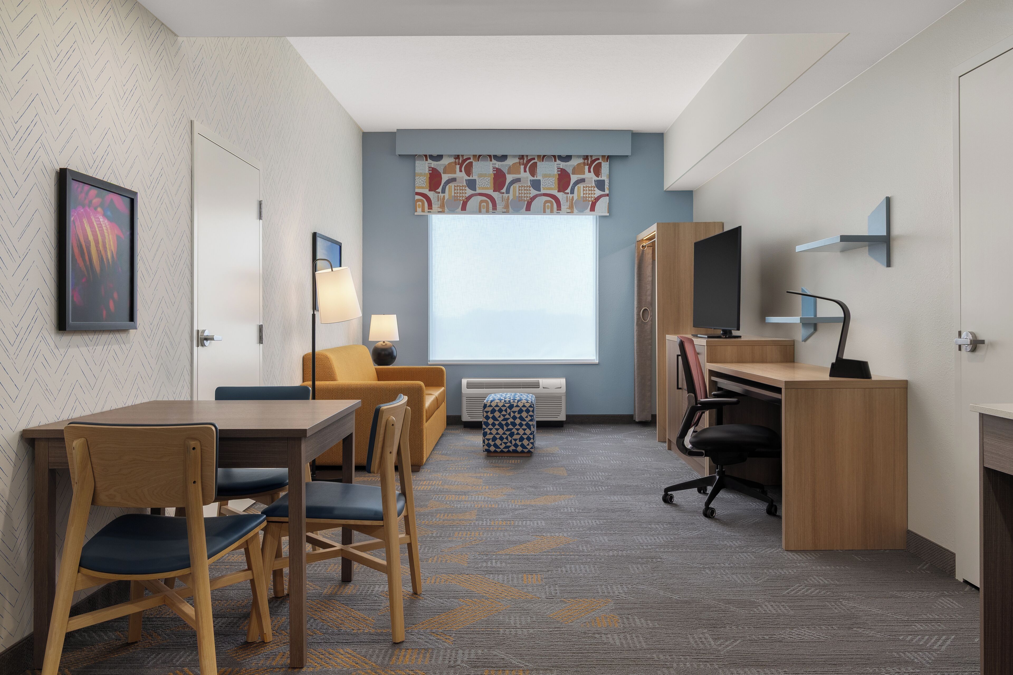 Quarto de hotel com escrivaninha, duas cadeiras, cama, janela com cortinas e televisão presa à parede.