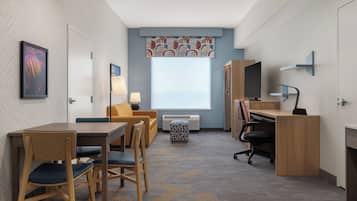 Quarto de hotel com escrivaninha, duas cadeiras, cama, janela com cortinas e televisão presa à parede.