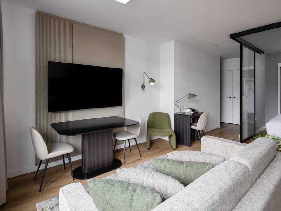 Uma sala de estar moderna com uma TV de tela plana, uma mesa de jantar, duas cadeiras, um sofá e uma pequena mesa lateral com um abajur.
