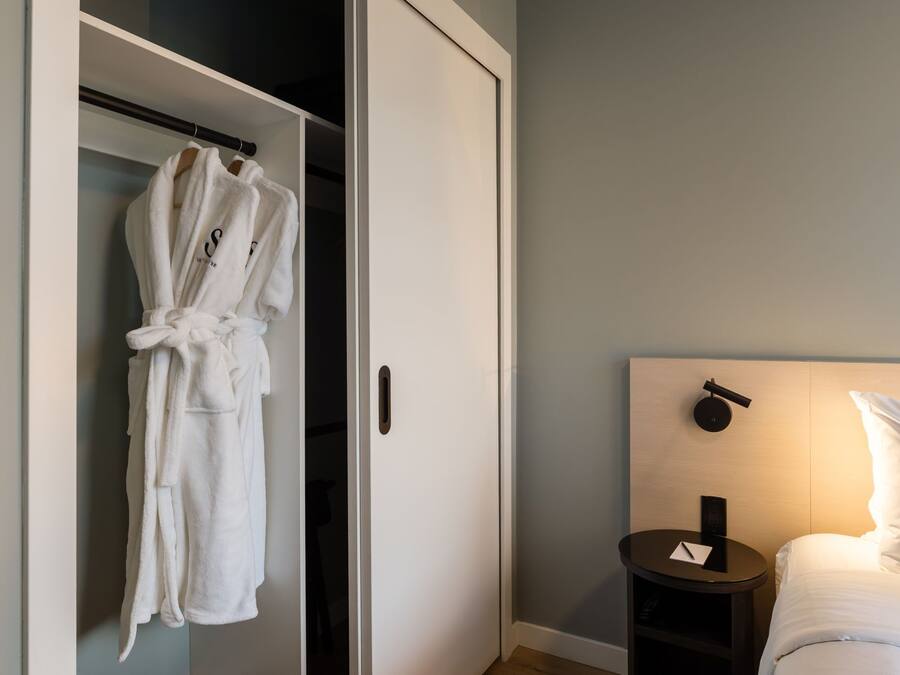 Quarto de hotel com cama branca, mesa de cabeceira com abajur e roupão pendurado na porta.